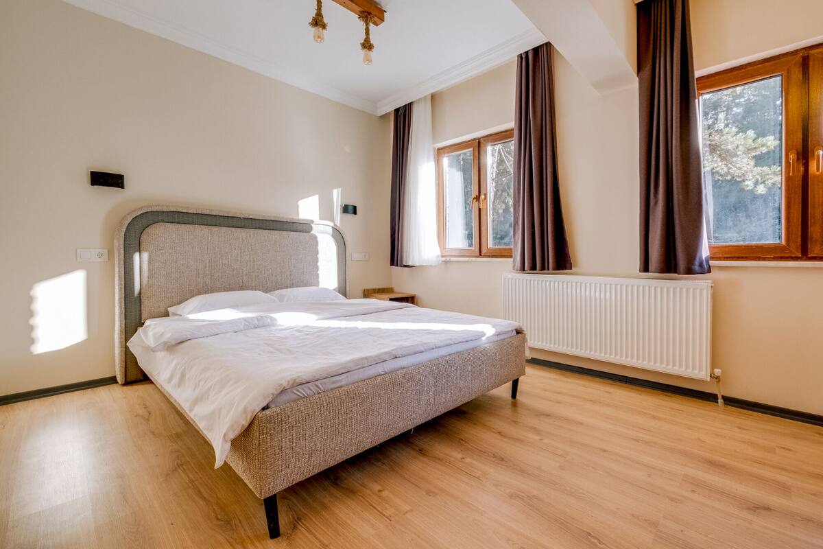 Quarto com cama, piso de madeira, janela com cortinas e radiador.