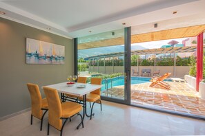 Uma sala de jantar moderna com uma mesa e cadeiras, um quadro na parede e vista para uma piscina e área externa com assentos.