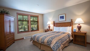 Quarto com uma cama grande, duas mesas de cabeceira com abajures, um guarda-roupa de madeira, uma janela com vista para árvores e um quadro na parede.
