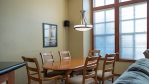 Uma área de refeições com uma mesa de madeira redonda, seis cadeiras, um lustre, um quadro na parede e uma janela com persianas.