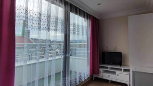 Quarto com uma janela grande, uma televisão sobre um suporte branco e uma varanda com guarda-corpo.