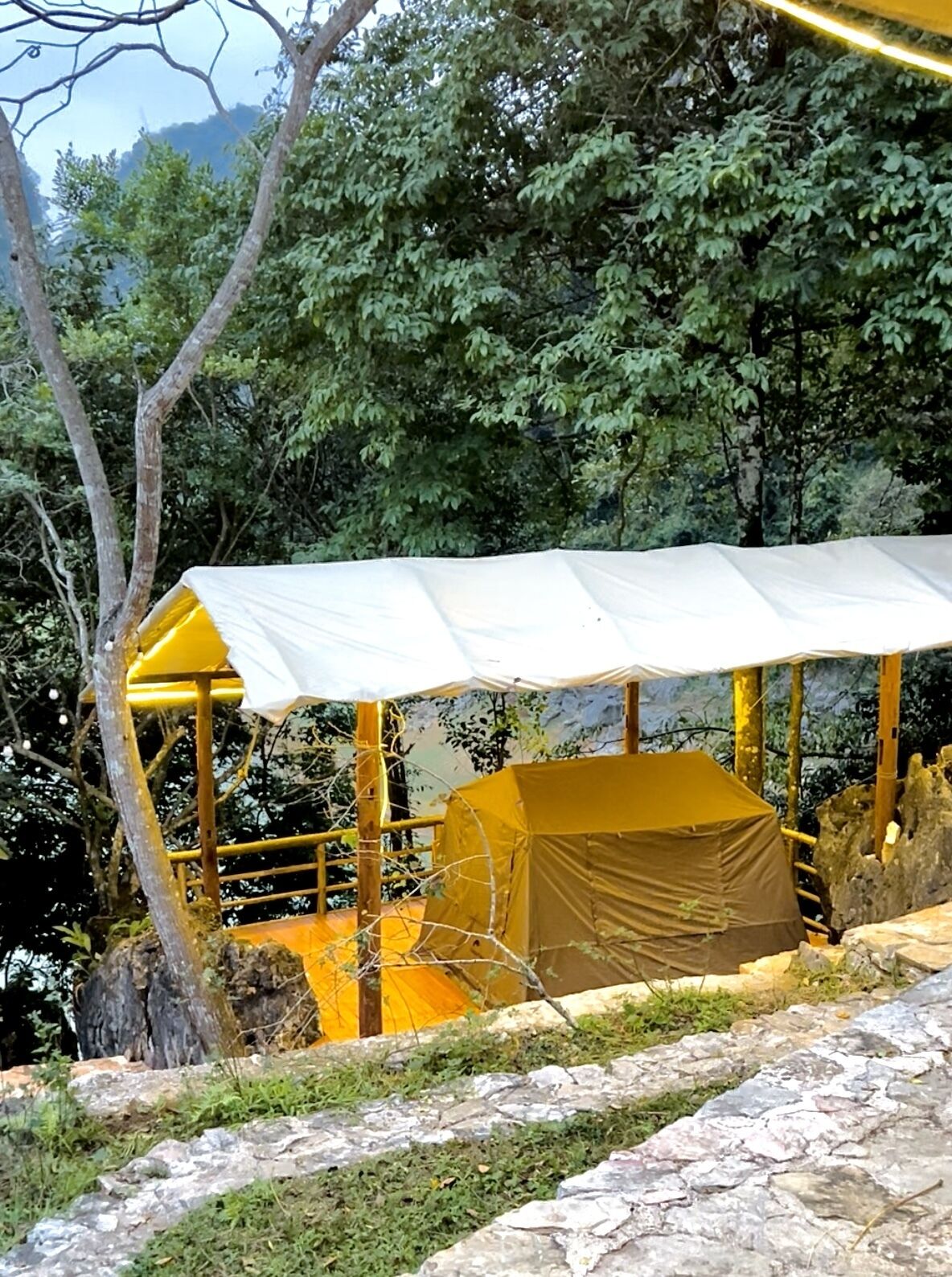 Uma tenda com teto branco e base amarela, rodeada por árvores e terreno rochoso.