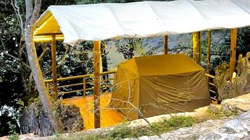 Uma tenda com teto branco e base amarela, rodeada por árvores e terreno rochoso.
