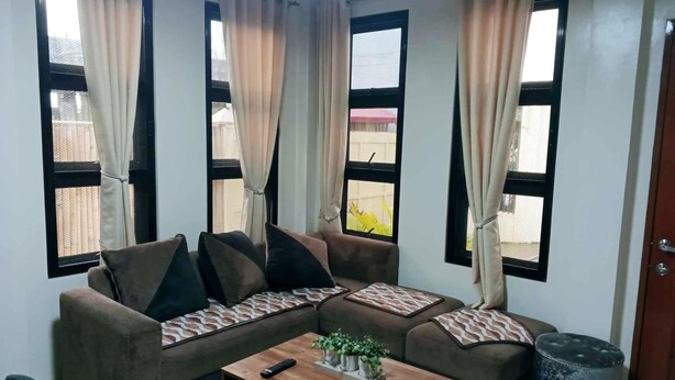 Sala de estar com sofá, mesa de centro e lixeira. Três janelas com cortinas.