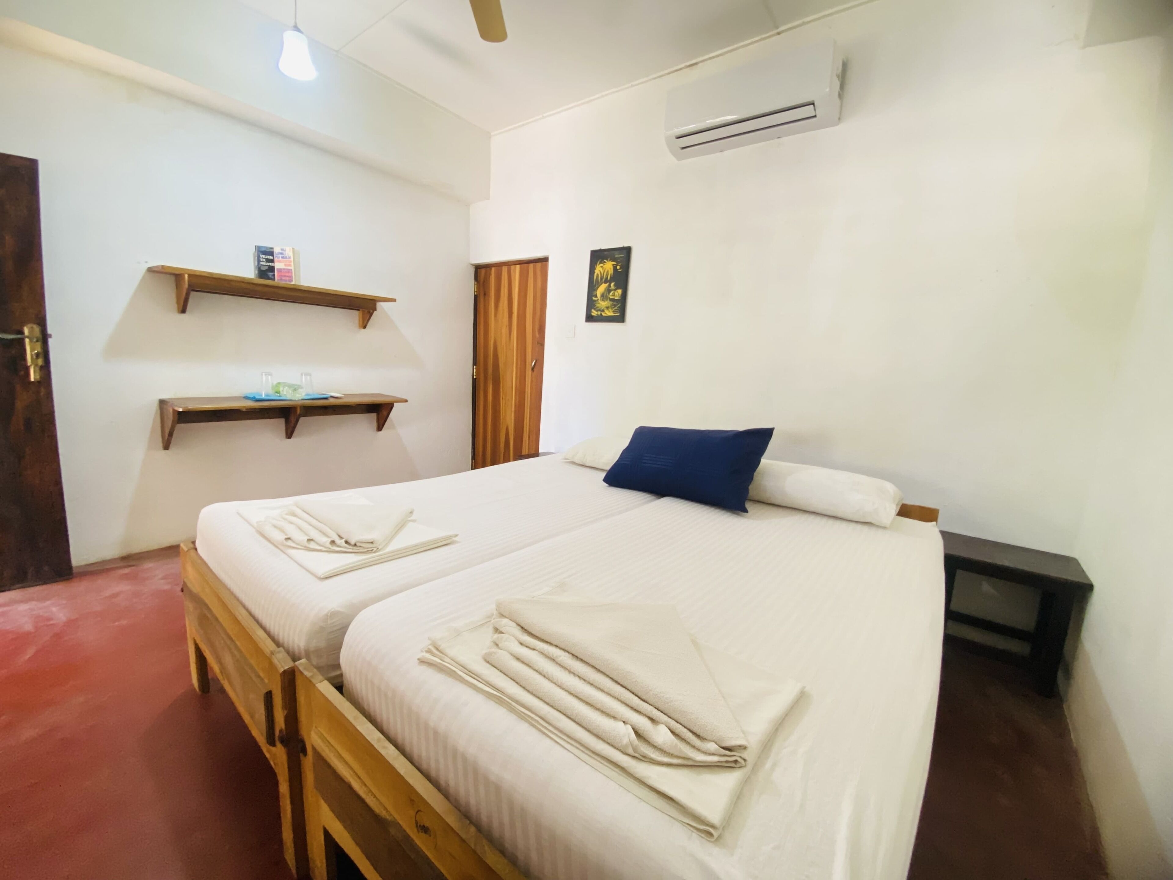 Quarto com duas camas, cabeceiras de madeira, prateleira de madeira com artigos de higiene pessoal, ar condicionado e uma porta.
