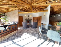 Um quarto espaçoso com piso de ladrilho de pedra, vigas de madeira no teto e uma lareira, além de uma área de jantar com mesa e cadeiras de vidro.
