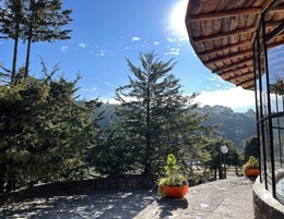 Um caminho de pedra leva a uma construção de madeira, com vista para árvores altas e montanhas.