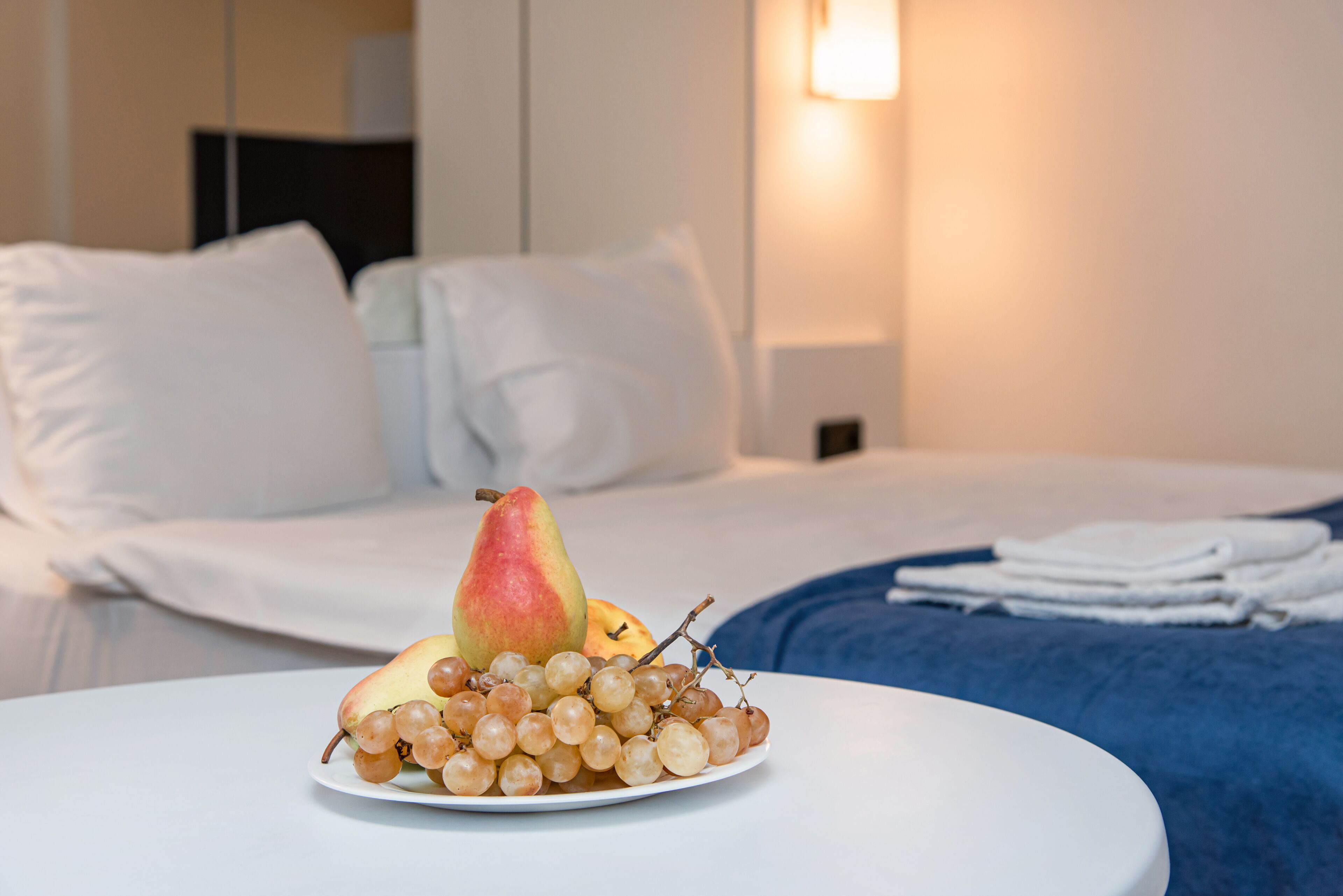 Uma cama com roupa de cama branca, uma mesa de cabeceira com uma travessa de frutas e um cobertor azul.