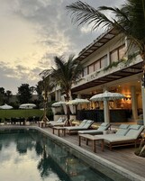 Uma área à beira da piscina com espreguiçadeiras e guarda-sóis, piscina e um edifício com varanda coberta.