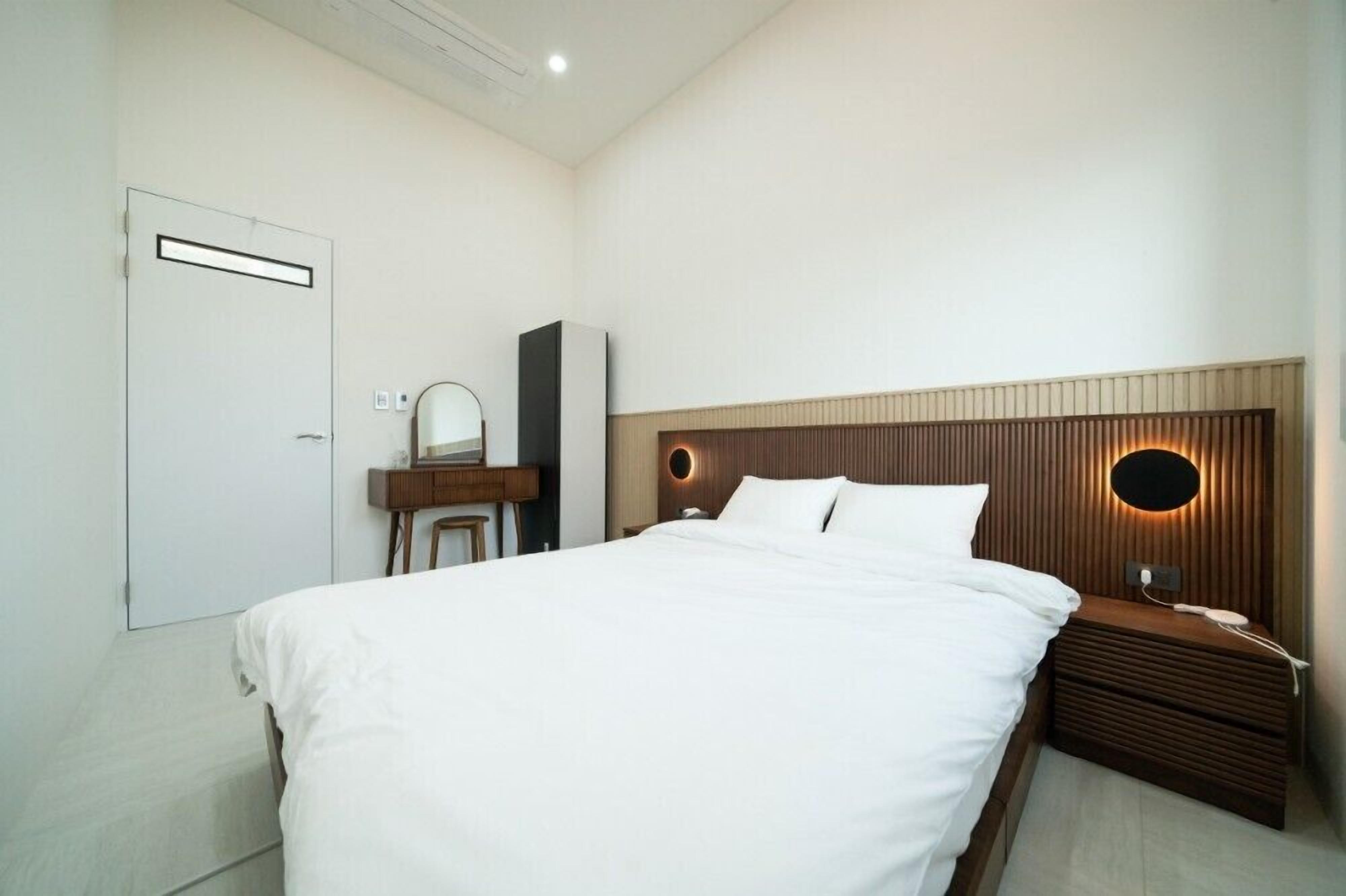 Quarto de hotel moderno com uma cama grande, uma mesa de cabeceira de madeira, uma escrivaninha com espelho e um candeeiro fixo na parede.