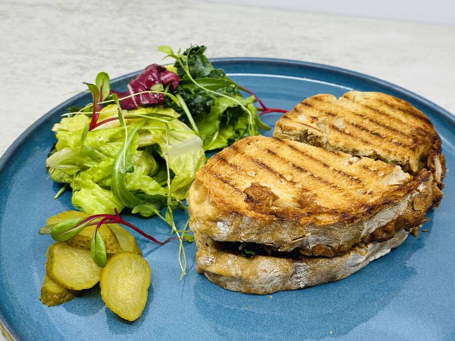 Sanduíche grelhado num prato azul, acompanhado de salada e picles.