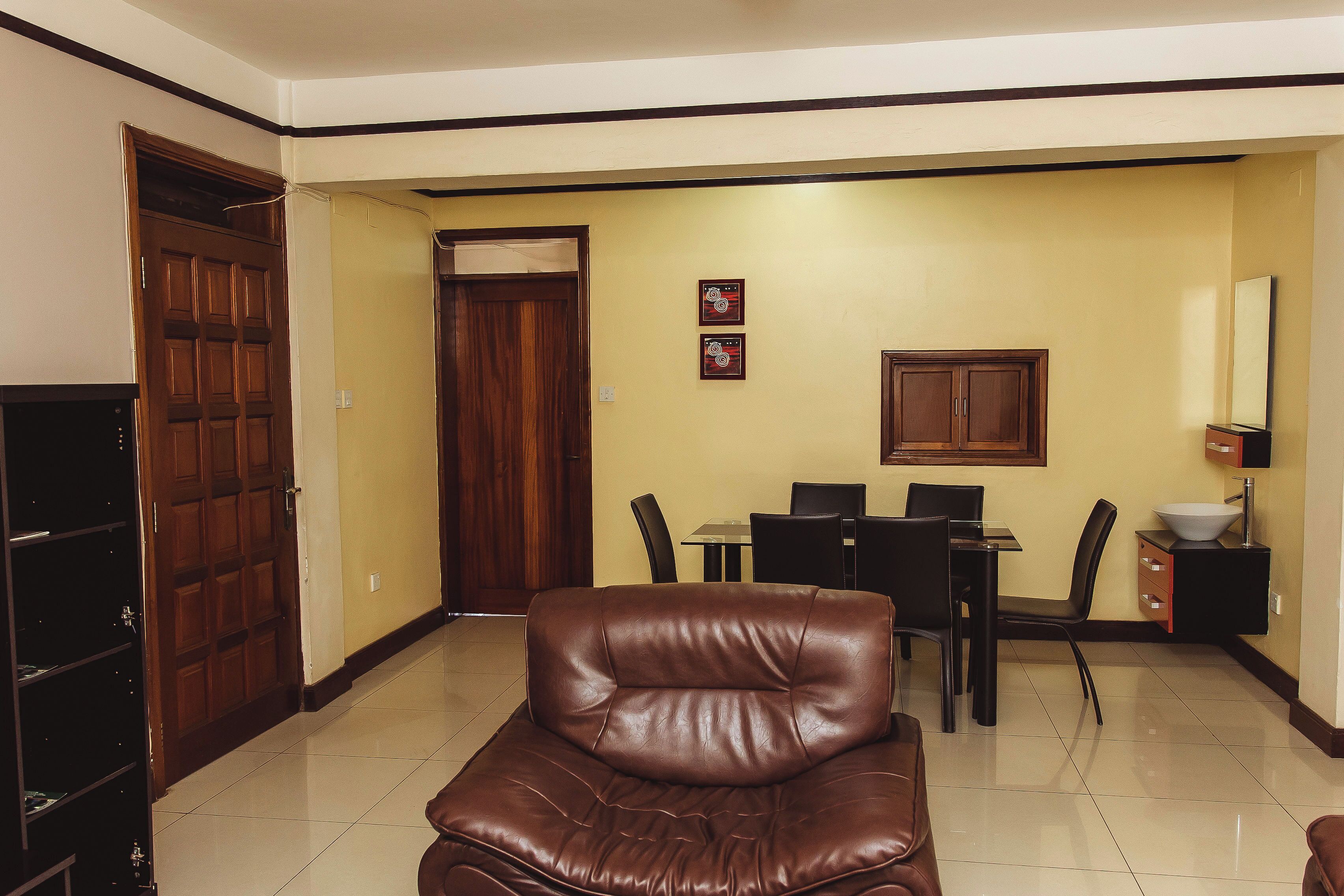 Uma sala de estar com um sofá de couro marrom, uma porta de madeira, uma mesa de jantar com cadeiras e um pequeno armário com espelho.