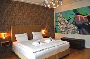 Quarto de hotel com uma cama grande, duas mesas de cabeceira, um criado-mudo, um banco de madeira e uma pintura abstrata colorida na parede.