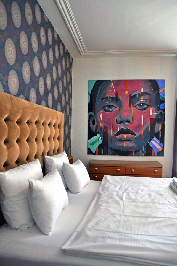 Quarto com cabeceira estofada, roupa de cama branca, cómoda de madeira e um grande quadro abstrato na parede.