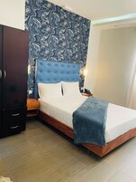 Uma cama bem arrumada com lençóis brancos e cabeceira azul, uma mesa de cabeceira de madeira com um telefone, e um guarda-roupa escuro.