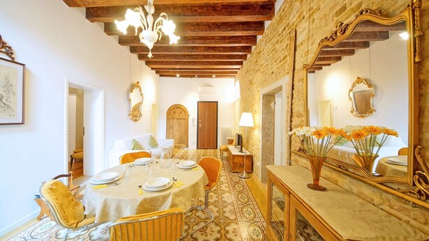 Uma sala de jantar com uma mesa redonda posta para quatro pessoas, um candelabro e um espelho com moldura dourada.