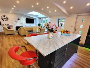 Cozinha moderna com bancada de mármore, banquetas vermelhas, uma ilha central com um vaso de flores e uma área de refeições com sofá e cadeiras.