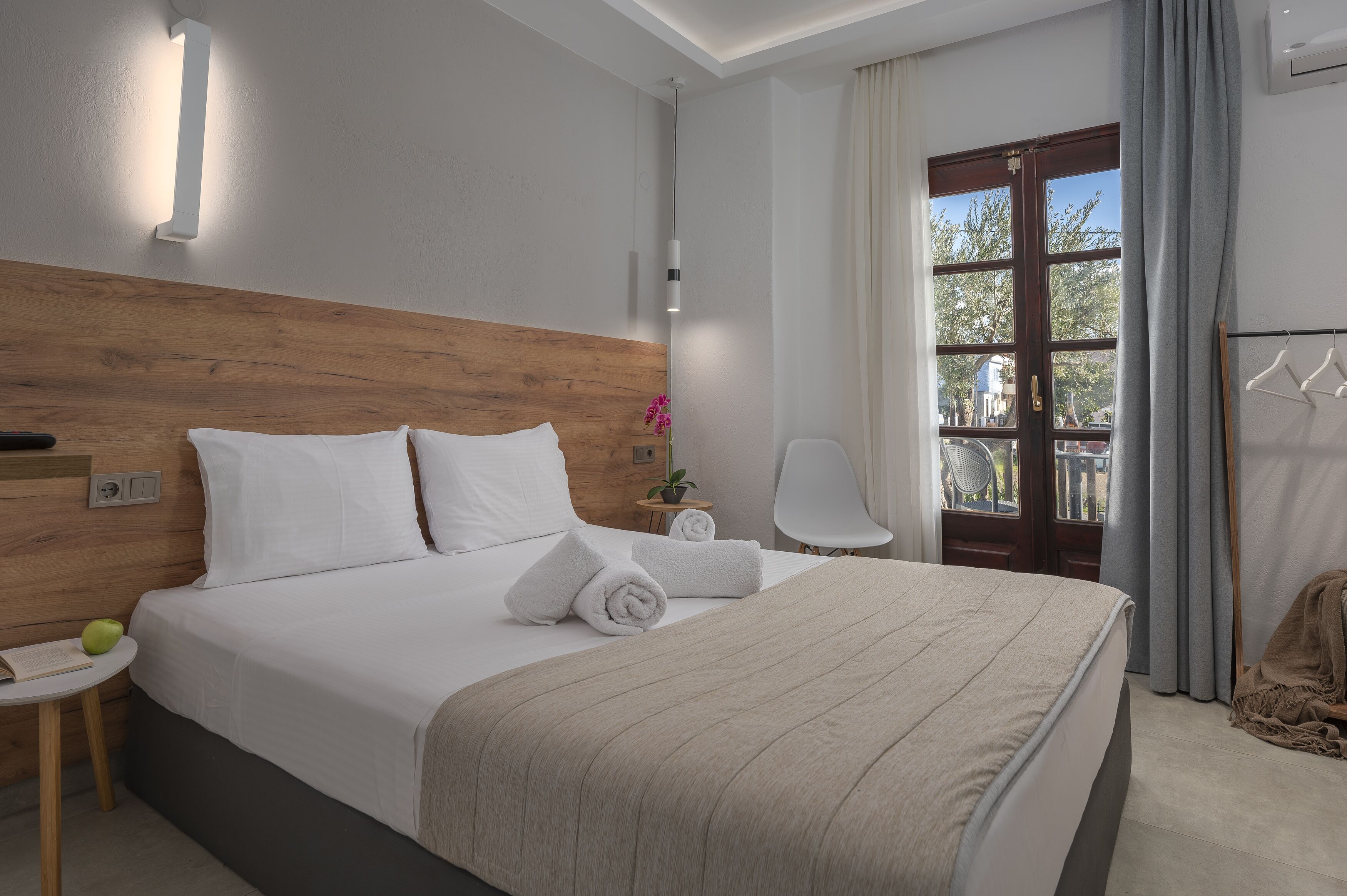 Quarto de hotel com cabeceira de madeira, cama com lençóis brancos, uma pequena mesa de cabeceira com um livro e uma fruta, uma cadeira e vista para uma varanda.