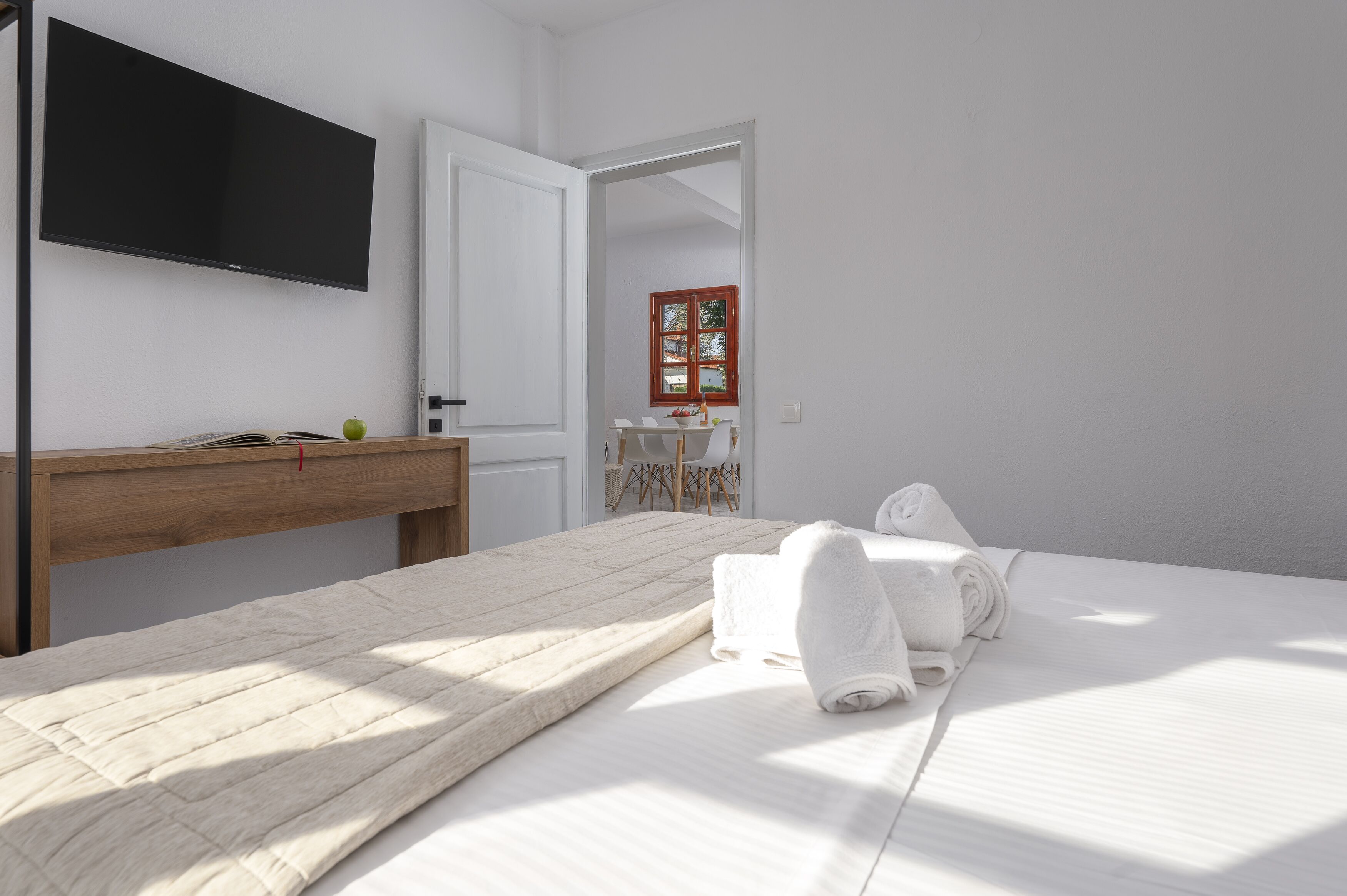 Quarto de hotel com cama, televisão de tela plana na parede, cabeceira de madeira e uma pequena mesa com cadeiras ao fundo.