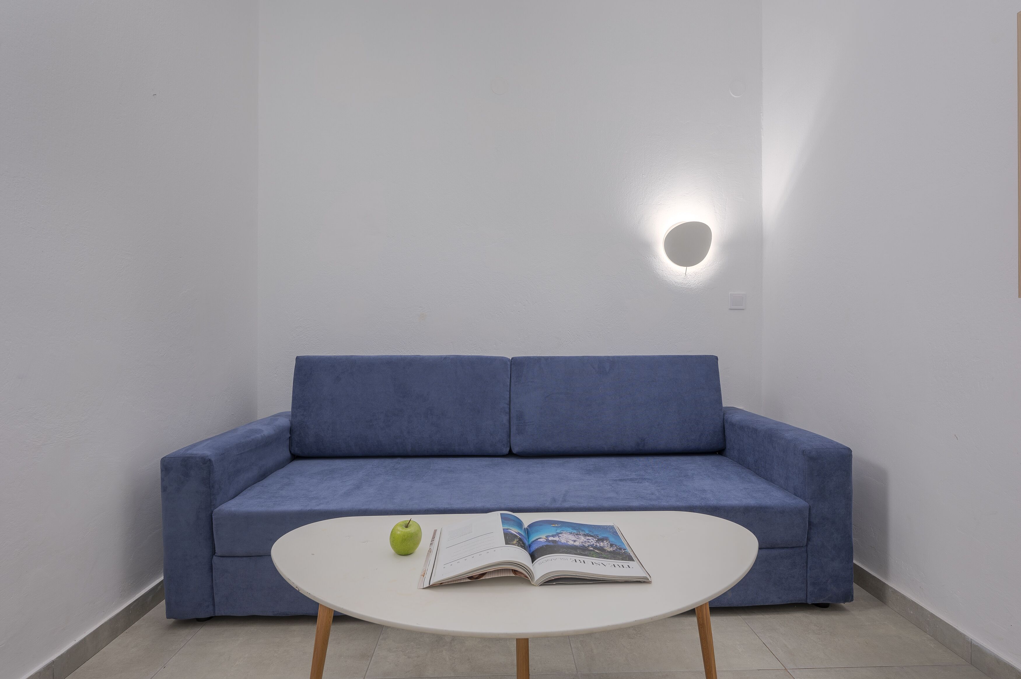 Um sofá azul com uma mesa branca na frente, uma maçã verde e uma revista aberta.