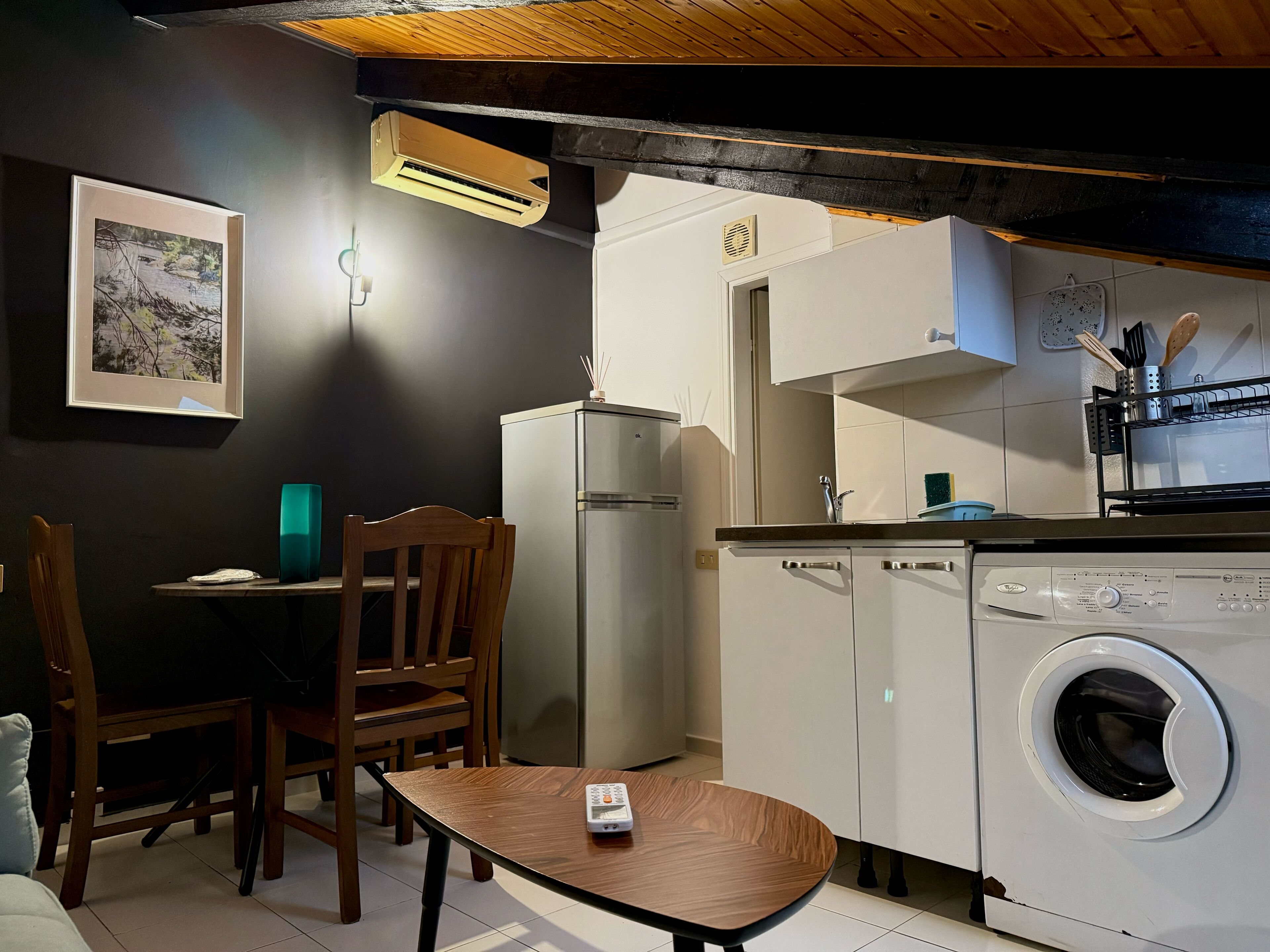 Cozinha compacta com máquina de lavar, geladeira, mesa de jantar com cadeiras e um quadro na parede.