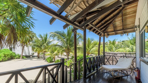 Um deck de madeira com telhado de palha, uma cadeira estofada e vista para palmeiras e uma praia.