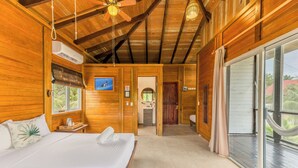 Quarto de cabana de madeira com cama, ventilador de teto, janela com persianas e porta para um banheiro.