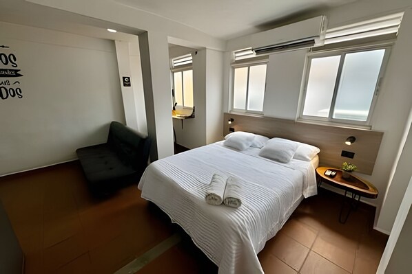 Quarto de hotel com cama, uma cadeira, mesa de cabeceira com abajur e janela.