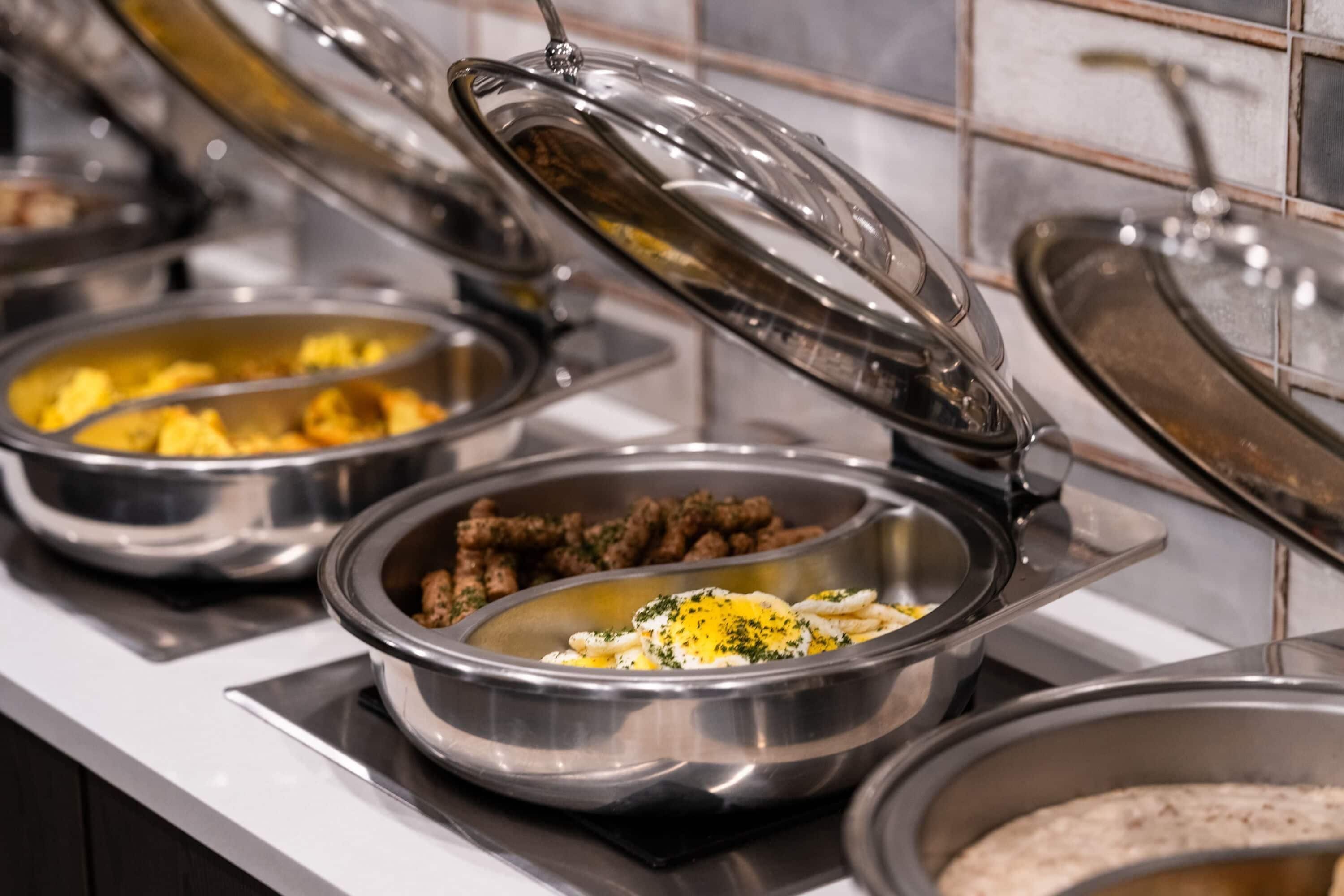 Buffet com diversos alimentos em recipientes de metal, alguns com tampas.