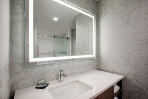 Casa de banho com parede em padrão marmorizado, lavatório branco, cabine de duche em vidro e espelho com moldura iluminada.
