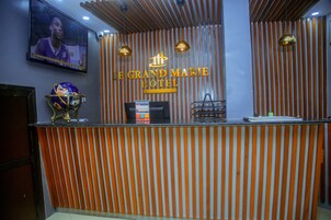 Uma receção com painel de madeira riscada, uma televisão montada acima e uma placa a indicar "Le Grand Marie Hotel".