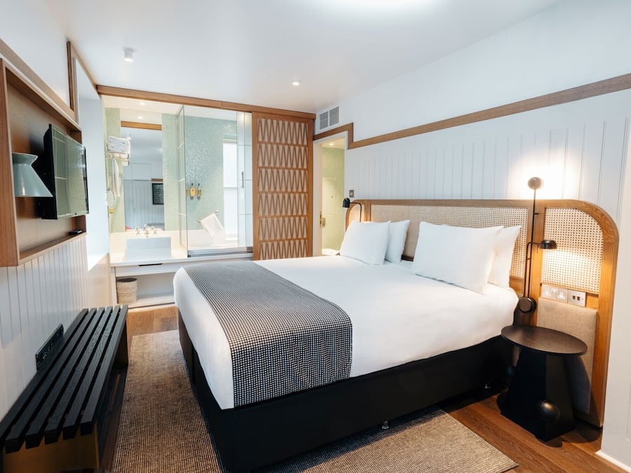 Quarto moderno com uma cama grande, uma mesa de cabeceira, uma luminária e vista para o banheiro.