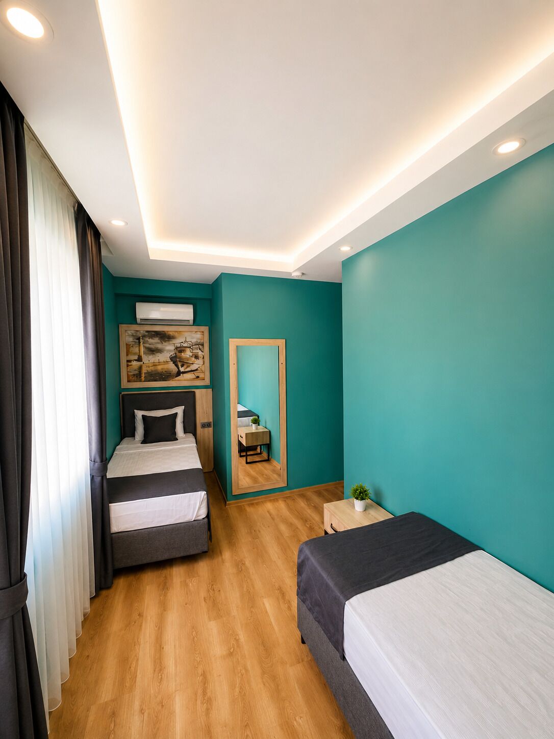 Quarto de hotel com duas camas, paredes verde-água, piso de madeira, um espelho grande e um aparelho de ar condicionado.