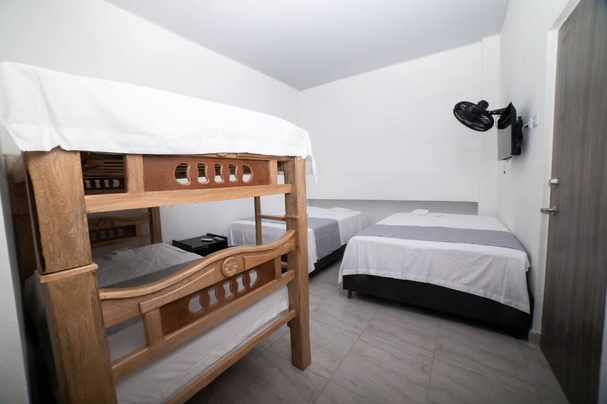 Quarto com beliche de madeira, duas camas simples, ventoinha na parede e uma porta.