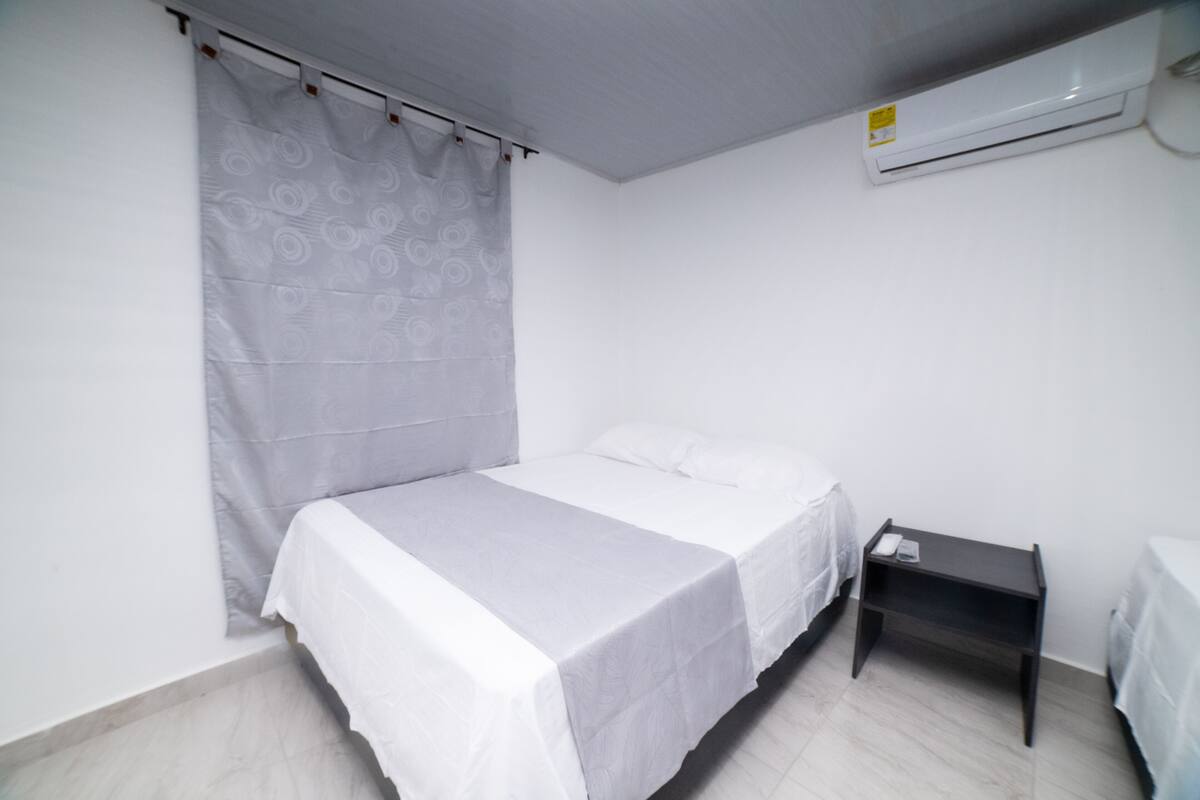 Uma cama de solteiro com um cobre-leito branco num quarto com paredes brancas e chão branco. Há uma pequena mesa de cabeceira com uma lâmpada e uma tomada. Um aparelho de ar condicionado está fixado na parede acima da cama.