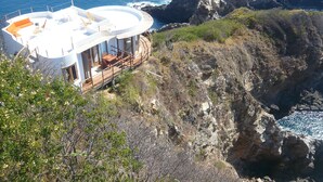 Uma casa à beira-mar com um deck circular, com vista para o mar e falésias rochosas.