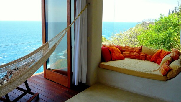 Uma cadeira rede em deck de madeira com vista para o mar, cama com roupa de cama laranja e bege, e vista para o oceano e vegetação.
