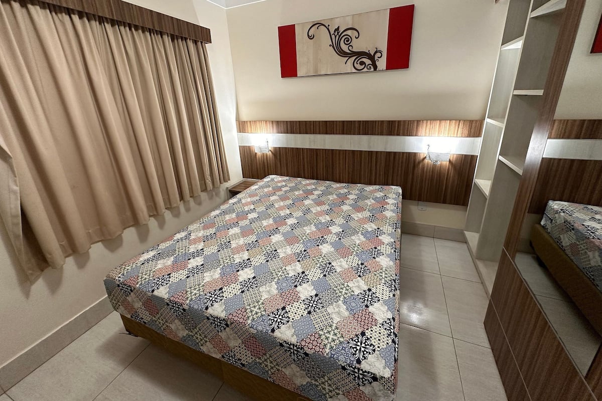 Quarto com cama, cortinas, luminária na parede e um objeto decorativo sobre a cama.