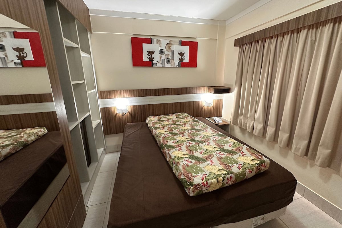 Quarto de hotel com cama, mesa de cabeceira, luminária, prateleira, janela com cortinas e uma parede com obras de arte.