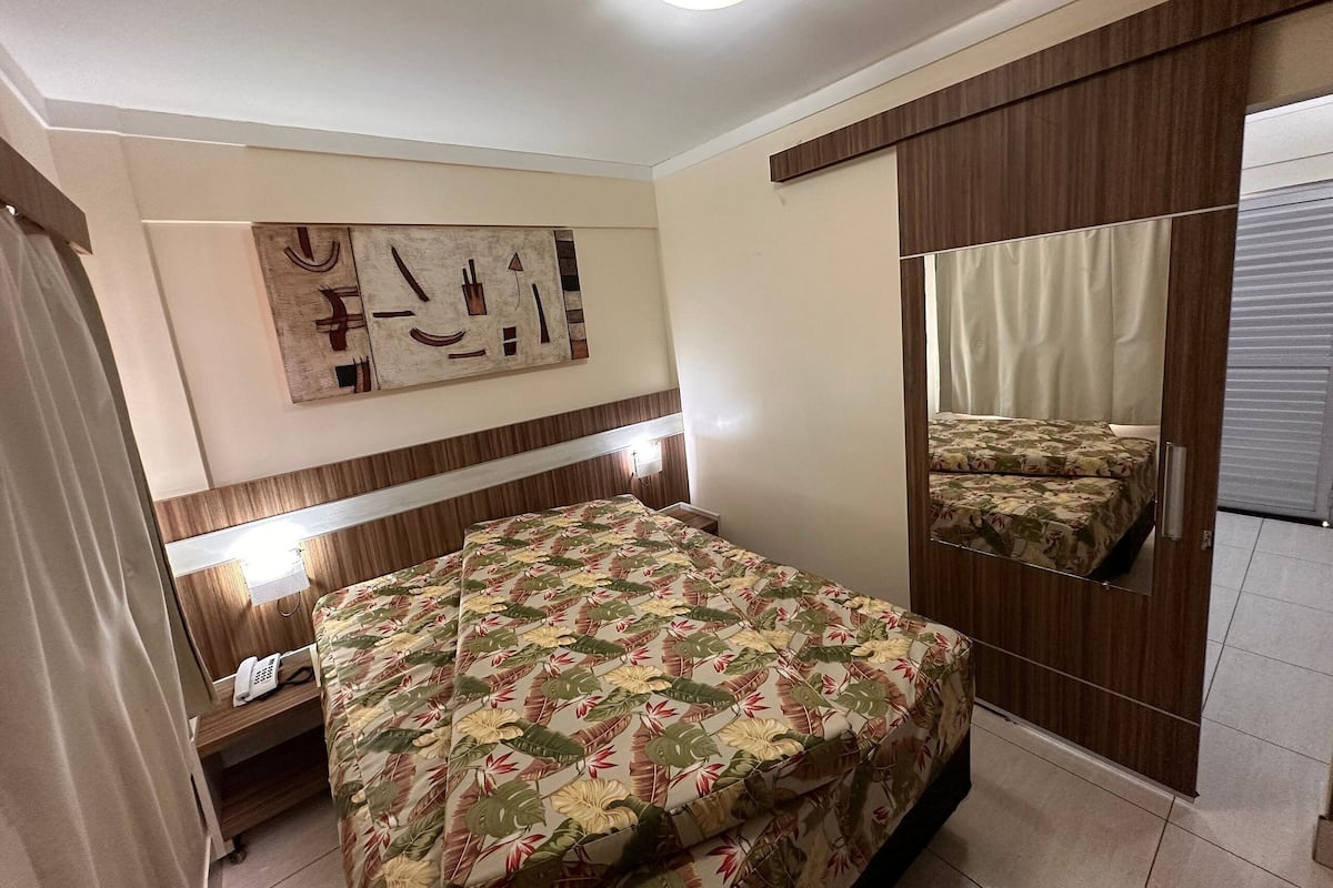 Quarto com cama, telefone de apoio, espelho e uma obra de arte na parede.