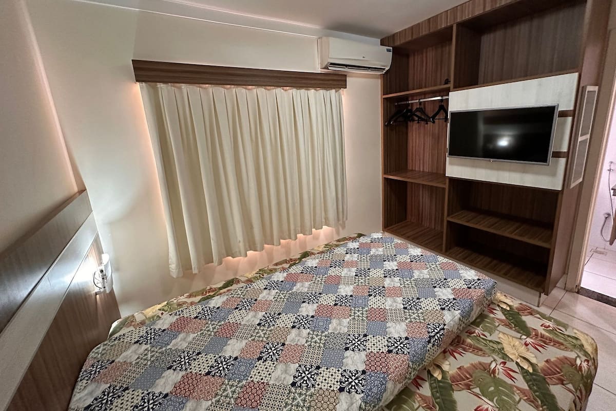 Quarto com cama, televisão presa a uma prateleira de madeira, ar-condicionado e janela com cortinas.