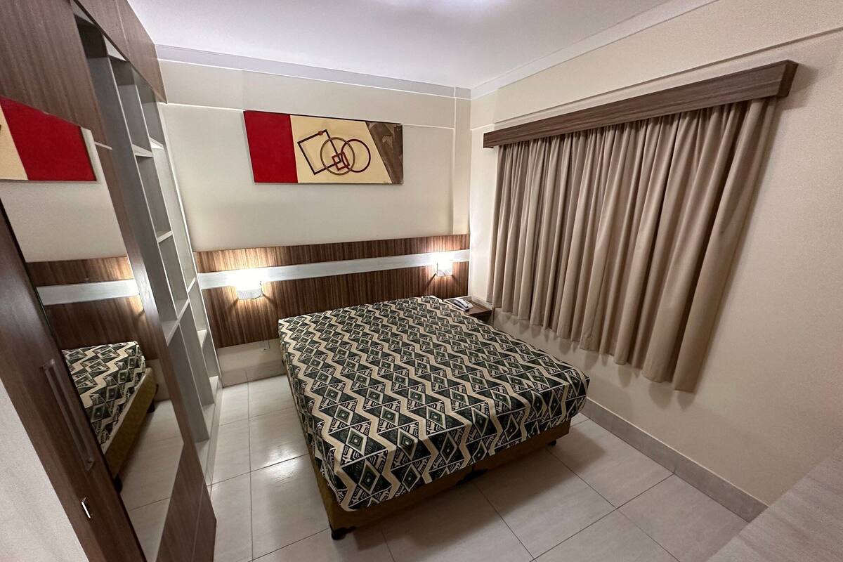 Quarto de hotel com cama, espelho, cabeceira, obra de arte pendurada na parede e janela com cortinas.