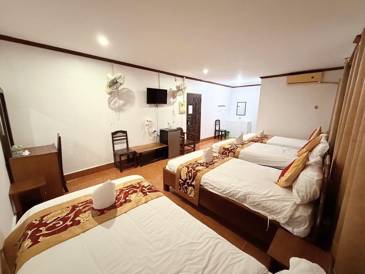 Quarto de hotel com quatro camas de solteiro, uma escrivaninha, uma cadeira, uma televisão e um ventilador fixado na parede.