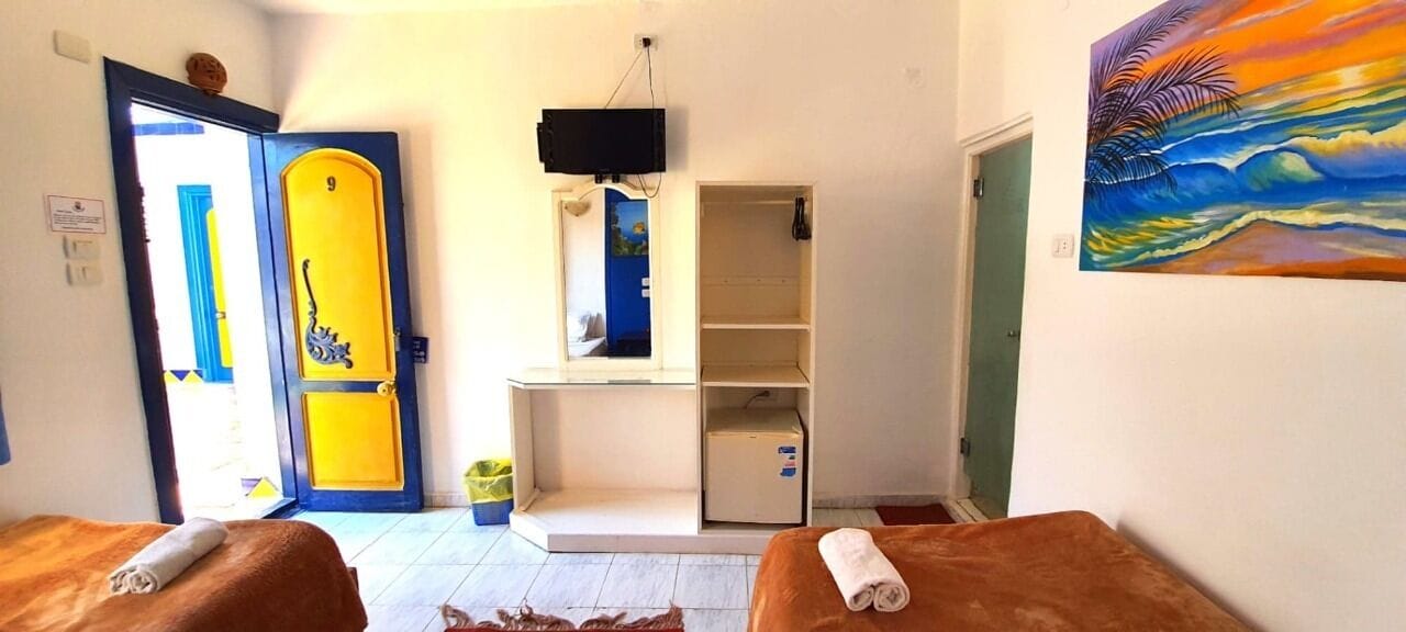 Quarto com duas camas, TV fixada na parede e um quadro de uma paisagem tropical.