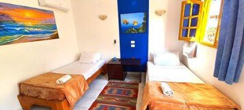 Quarto de hotel pequeno com duas camas de solteiro, uma mesinha, uma porta azul e um quadro de um pôr do sol na parede.