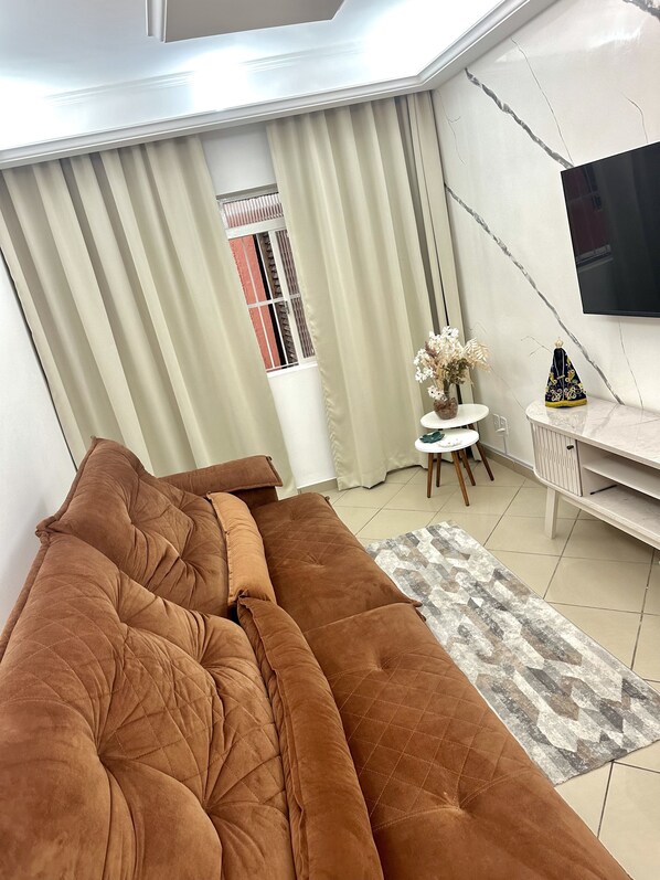 Uma sala de estar moderna com um sofá marrom grande, uma TV fixada na parede e uma mesa redonda pequena com um vaso de flores.
