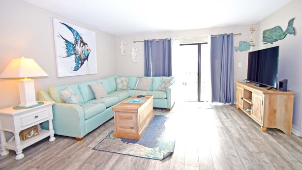 Uma sala de estar com um sofá azul claro, uma mesa de centro de madeira, uma TV em um suporte de madeira e um abajur sobre uma mesa de apoio branca.