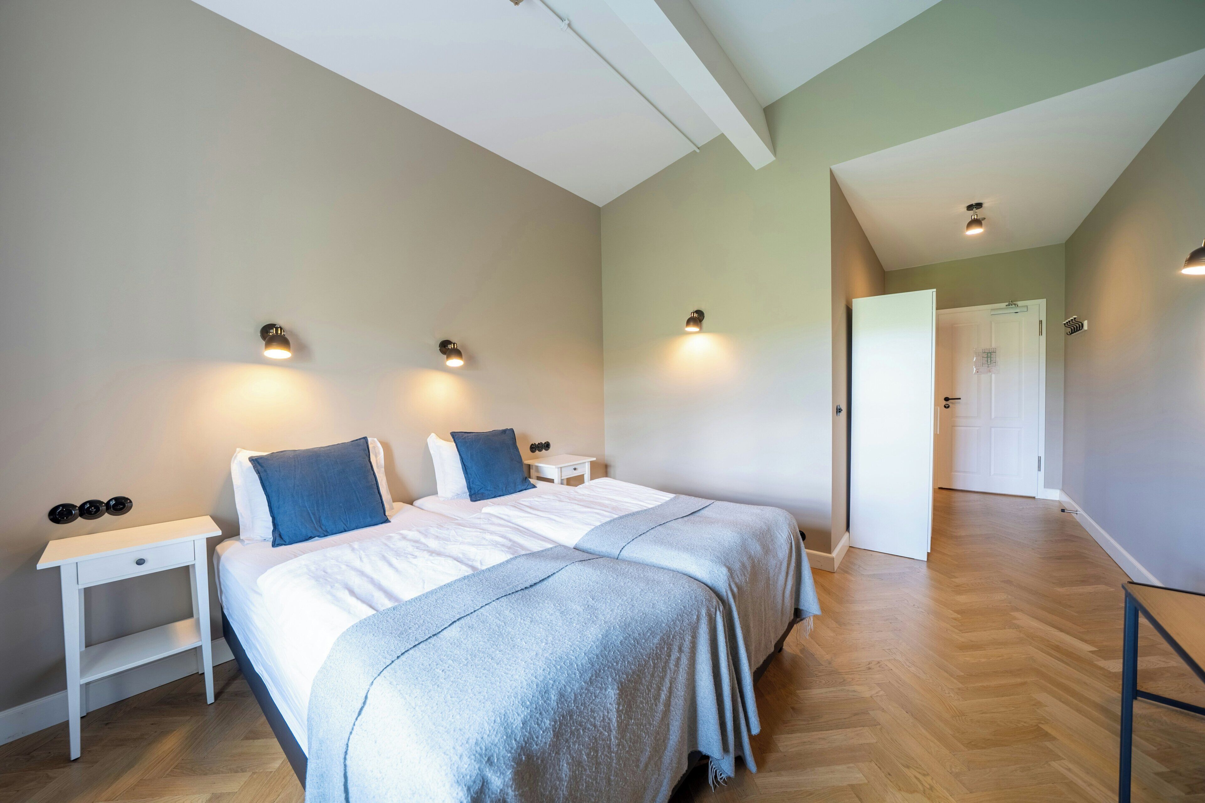 Quarto com duas camas, uma mesa de cabeceira branca, cômoda branca e piso de madeira.