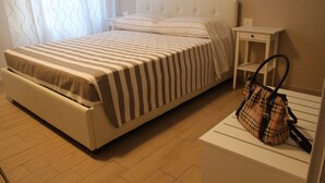 Cama bem arrumada com roupa de cama listrada, mesa de cabeceira com abajur e uma bolsa em um suporte.