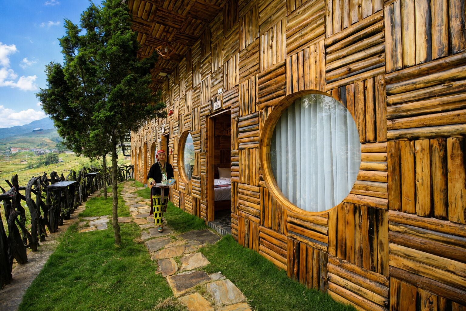 Fileira de edifícios em estilo de cabana de toras, com janelas redondas, um caminho de pedra e uma pessoa vestida com traje tradicional.
